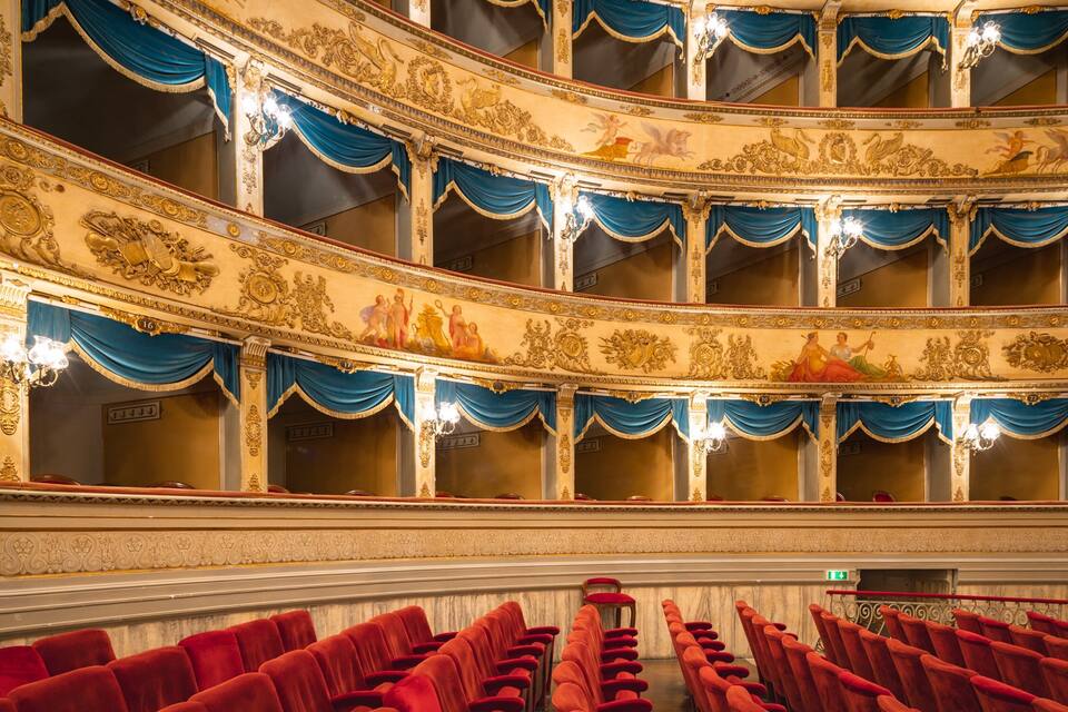Um teatro imponente com decoração dourada e azul ricamente detalhada, assentos de veludo vermelho e um balcão com um mural.