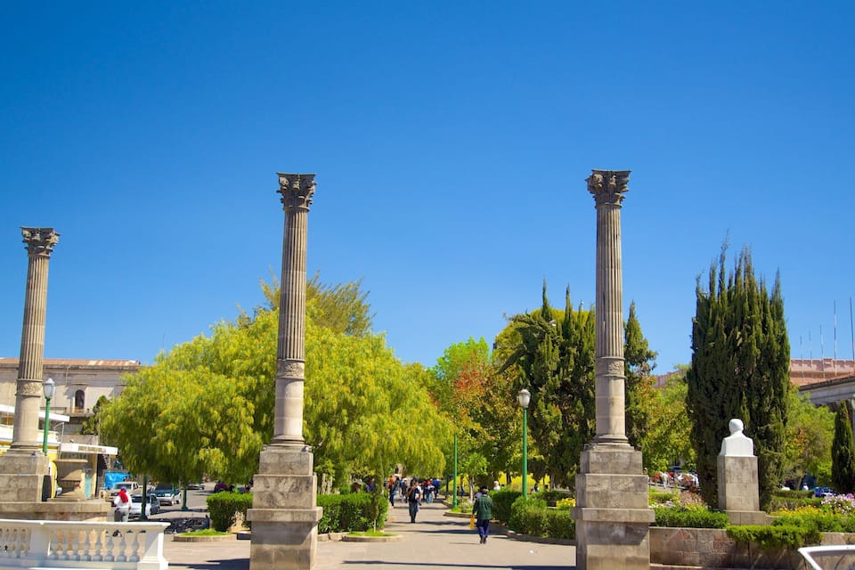Uma praça com quatro colunas clássicas, cercada por árvores e um céu azul límpido.