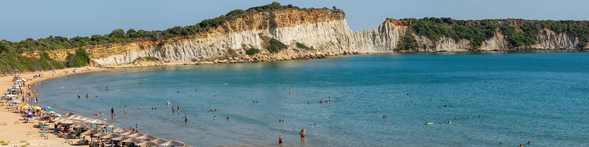 Uma praia com águas azul-claras, pessoas nadando e tomando sol, e falésias ao fundo.