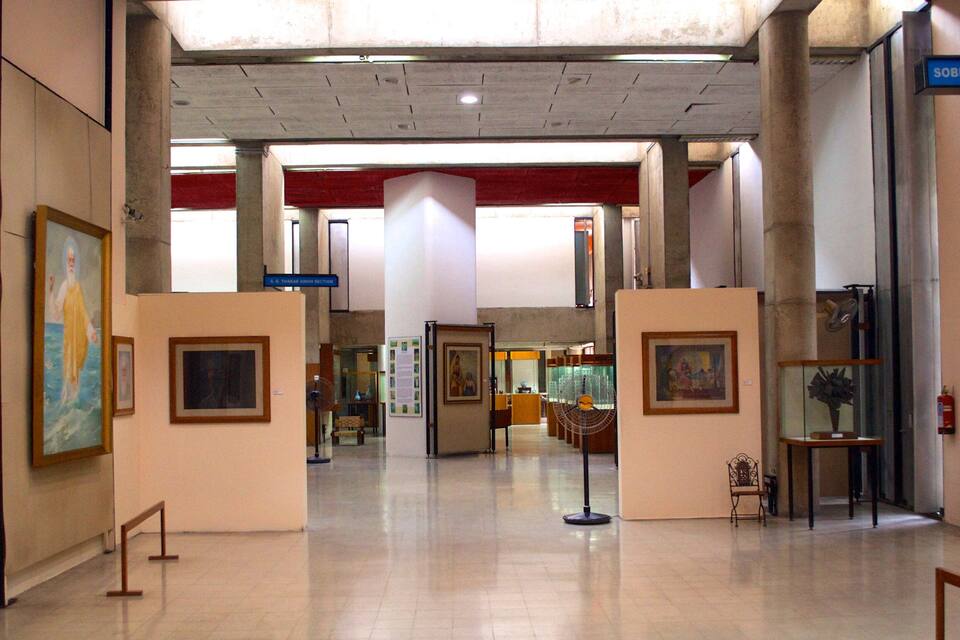Uma galeria de arte com pinturas expostas, teto com iluminação e uma placa que diz 'SOBHA SINGH SECTION'.