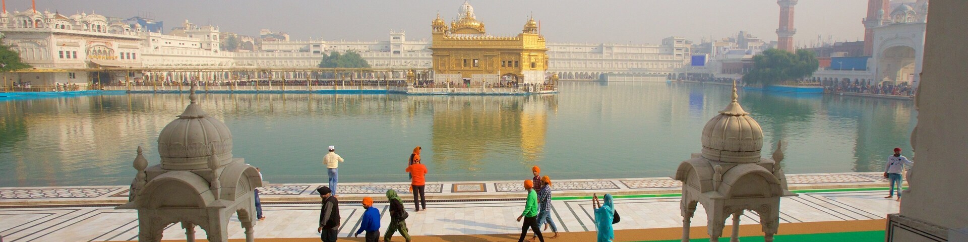 Visão do Templo Dourado refletido na água, rodeado por arquitetura tradicional e um grupo de pessoas caminhando em uma plataforma próxima.