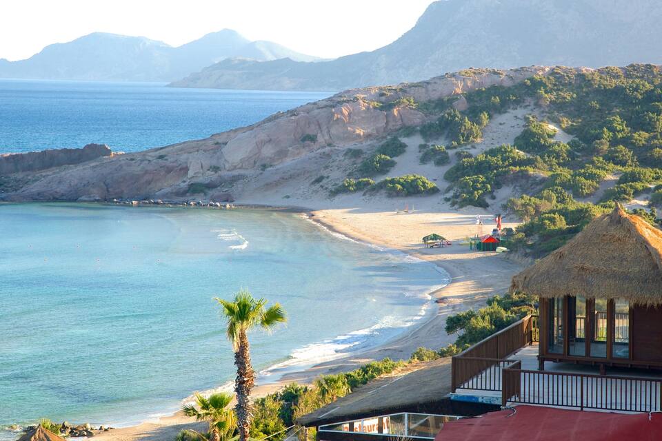 Paisagem costeira com praia, palmeiras, um edifício com telhado de palha e montanhas ao fundo.