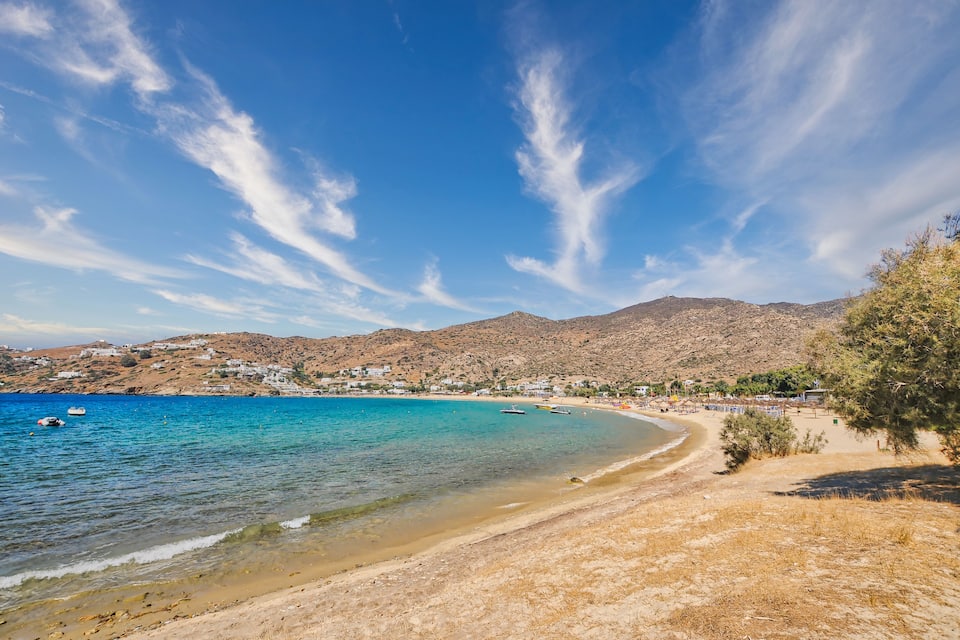 Paisagem costeira com céu azul límpido, praia arenosa e montanhas ao fundo.