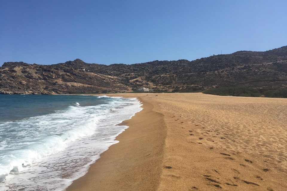 Uma praia arenosa com ondas, colinas rochosas e céu claro.