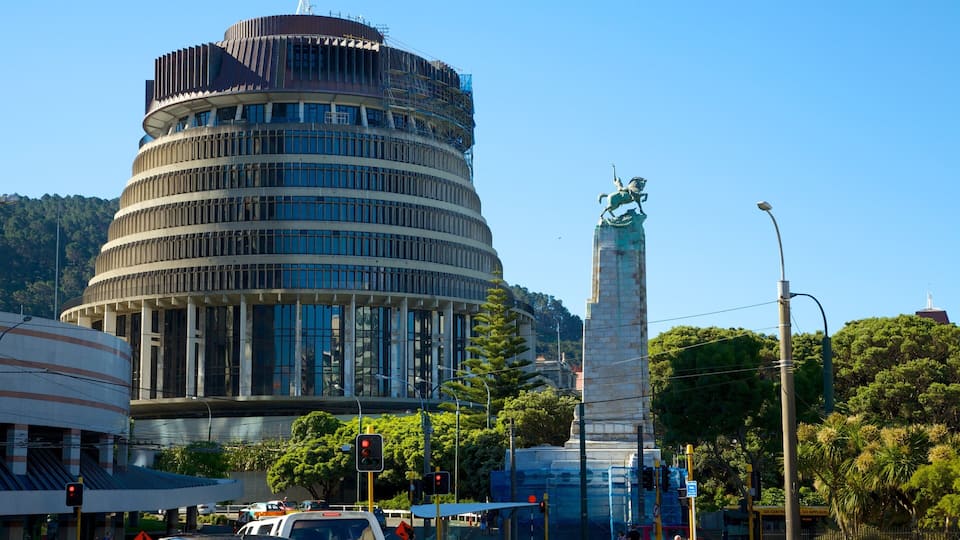 Um edifício moderno com design cilíndrico, um alto monumento com uma estátua, e um cruzamento movimentado com semáforos.