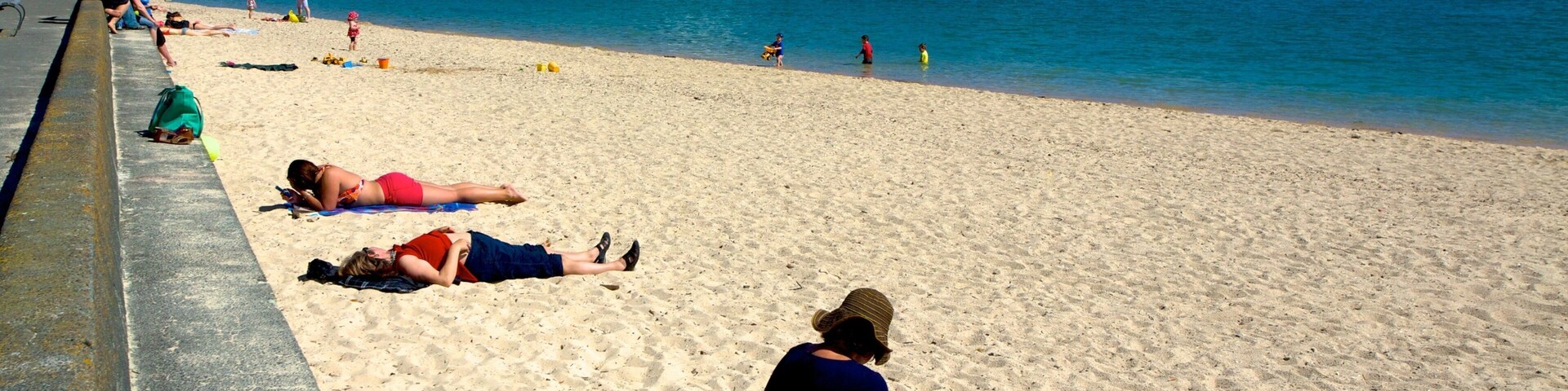Uma praia arenosa com pessoas tomando sol e um horizonte urbano ao fundo.
