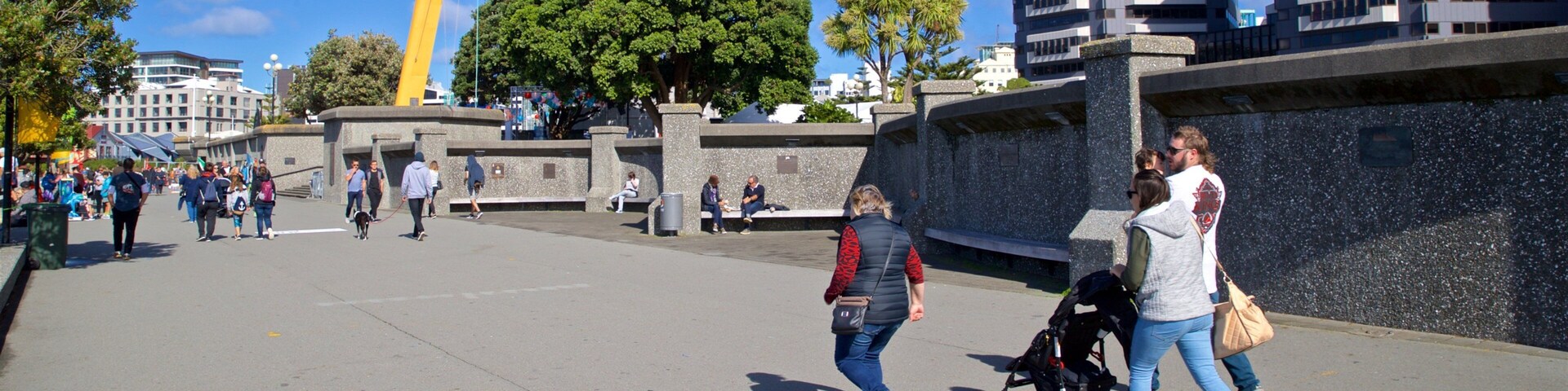 Uma praça pública com pessoas caminhando, uma torre amarela e edifícios modernos.