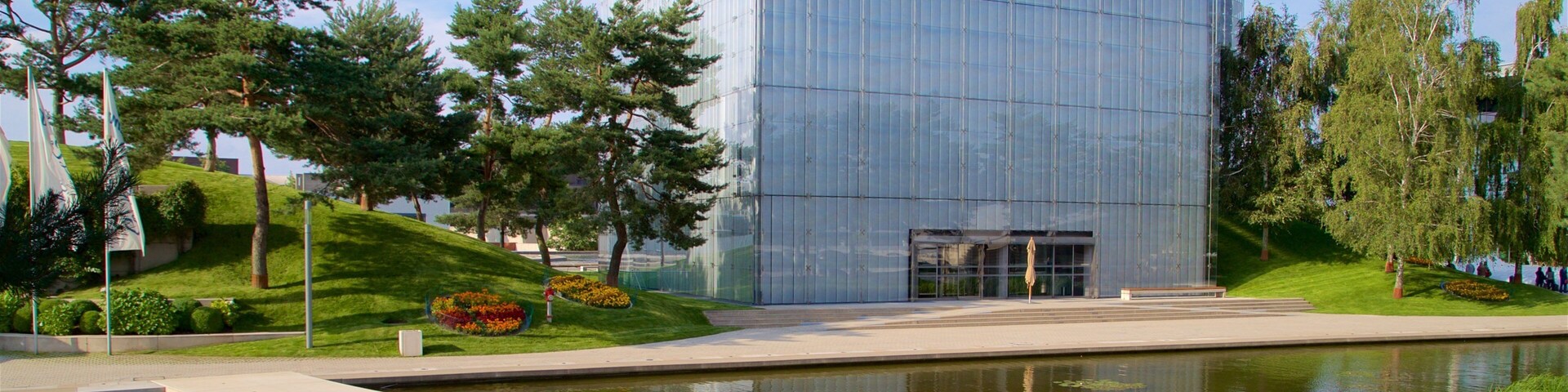 Um edifício moderno de vidro com fachada refletora, cercado por um gramado bem cuidado, um pequeno lago e algumas árvores.