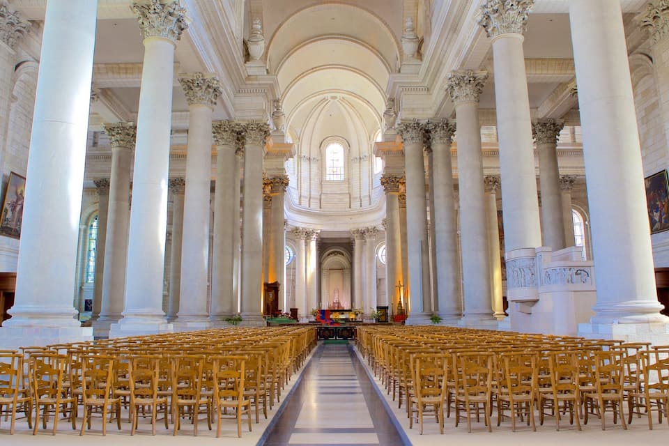 Um salão imponente com fileiras de cadeiras de madeira, colunas brancas e um corredor central que leva a um altar.