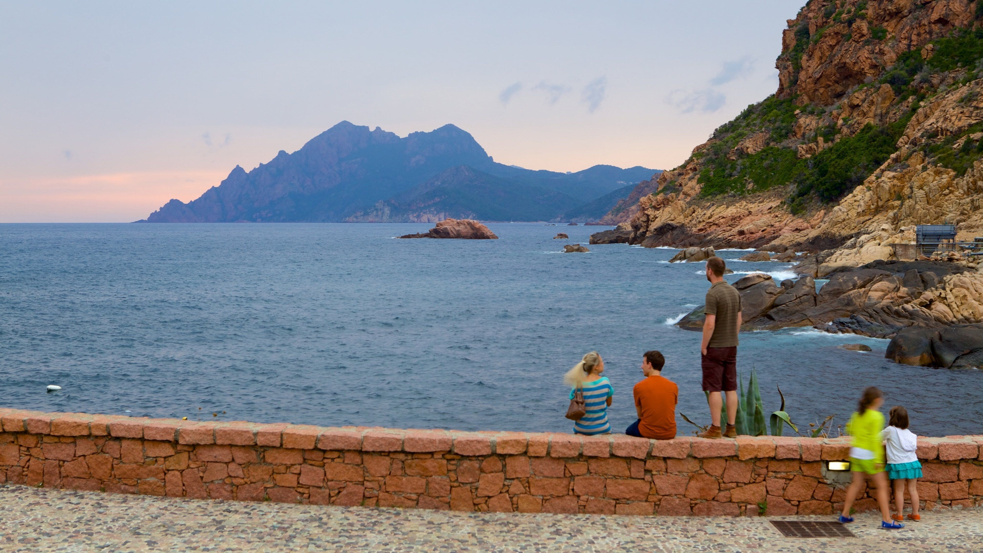 Uma paisagem costeira com um penhasco rochoso, um muro de pedra e um mar calmo.