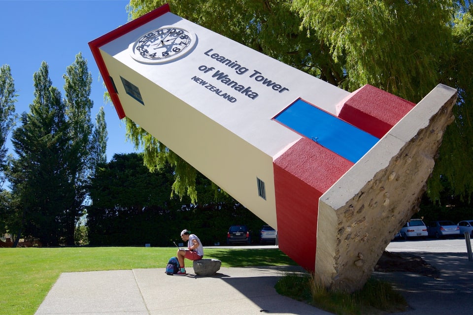 Um sinal inclinado para a Torre Inclinada de Wanaka, Nova Zelândia, com uma pessoa sentada por perto.