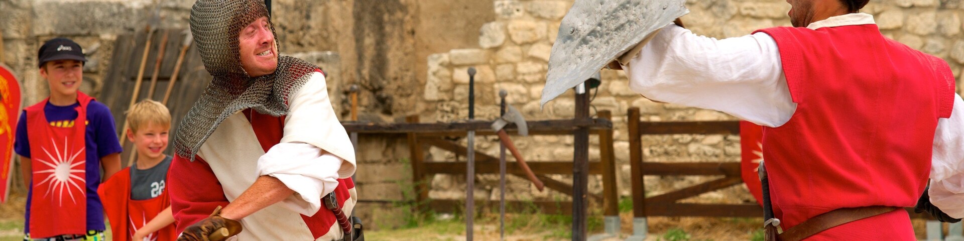 Reconstituição medieval com participantes usando cotas de malha e túnicas vermelhas, um muro de pedra e um portão de madeira ao fundo.