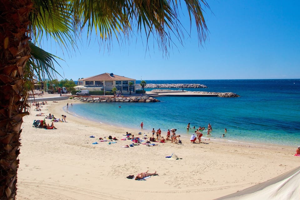 Uma praia com pessoas se banhando ao sol e nadando, um edifício e afloramentos rochosos na água.