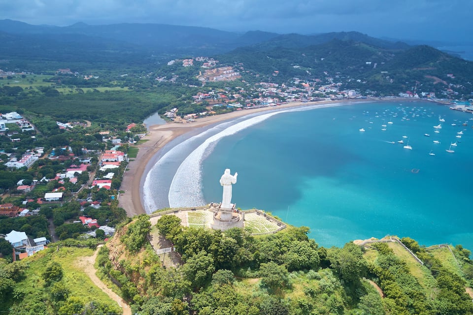 Uma vila costeira com uma estátua numa colina, com vista para uma praia e um porto de recreio.