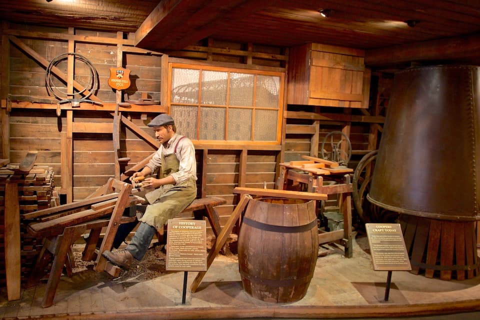 Instalação histórica de destilharia da Cooper's Guild de 1501, com barris de madeira, um alambique de cobre grande e um homem a operar uma prensa de madeira.