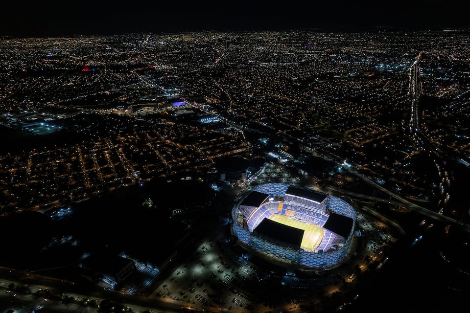 Vista aérea noturna de uma cidade, com um estádio em destaque, cercado por ruas e edifícios iluminados.