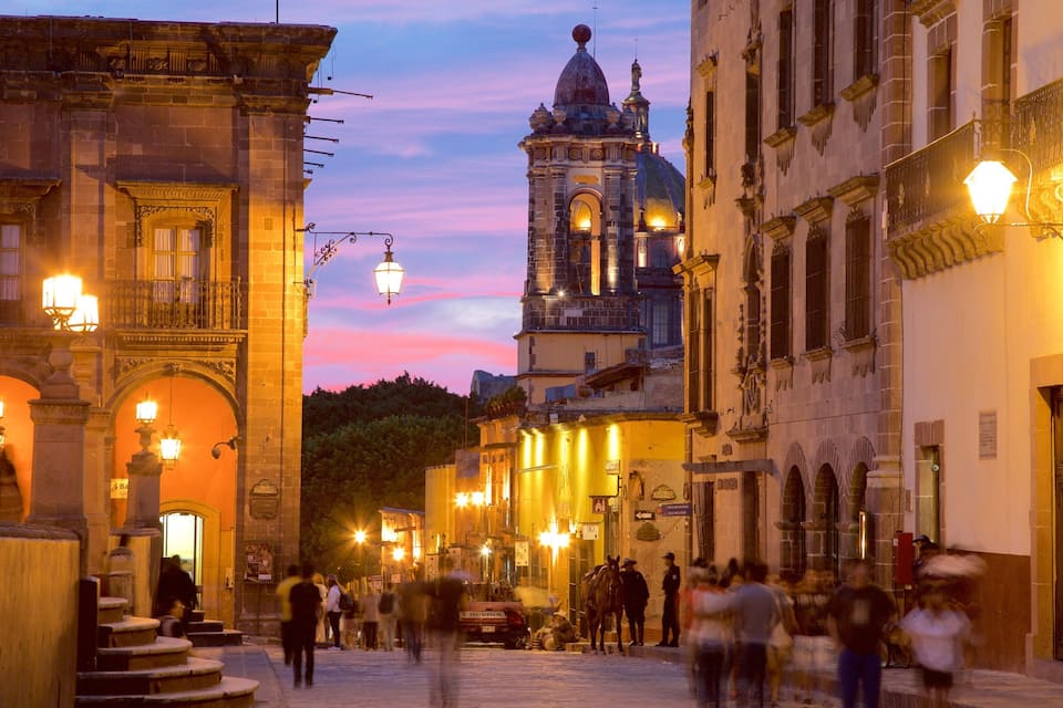 Uma praça histórica com edifícios ricamente decorados, uma torre de igreja imponente e pessoas caminhando.