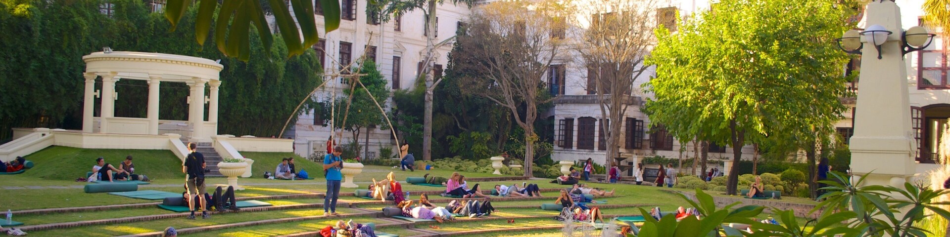 Um parque com pessoas relaxando em esteiras, um coreto e um edifício ao fundo.