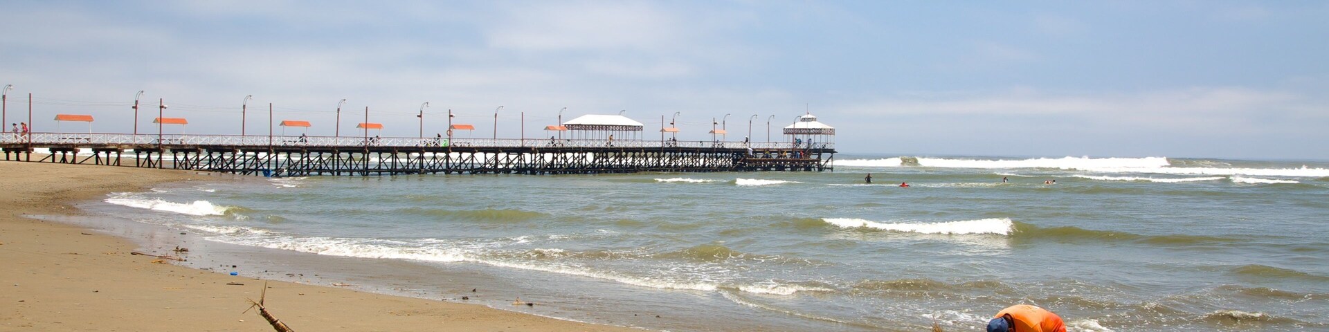 Uma praia com duas grandes embarcações de palha, um píer com guarda-sóis e ondas quebrando na costa.