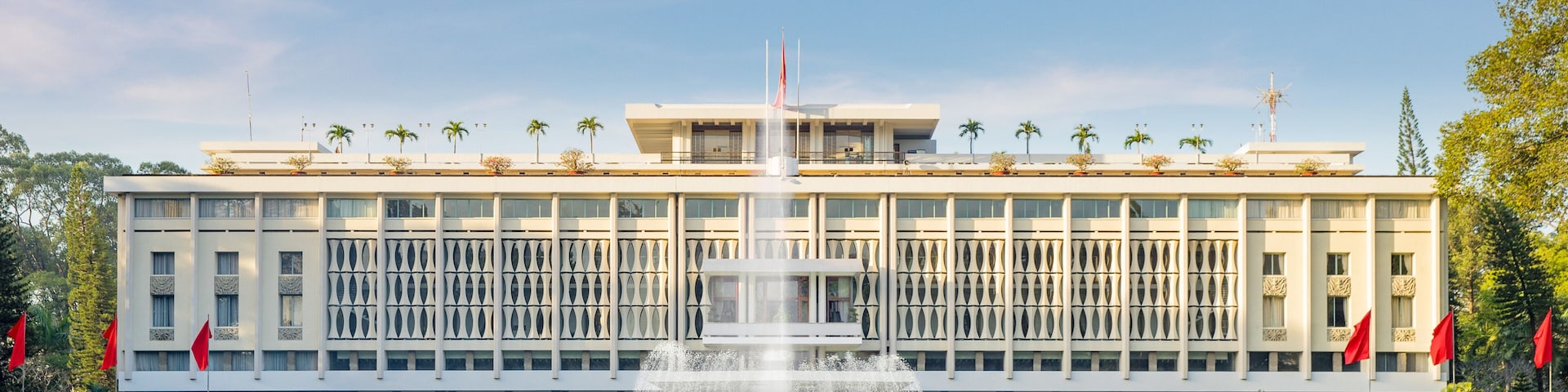 Um edifício grande com uma fonte central, rodeado por um jardim bem cuidado e palmeiras.