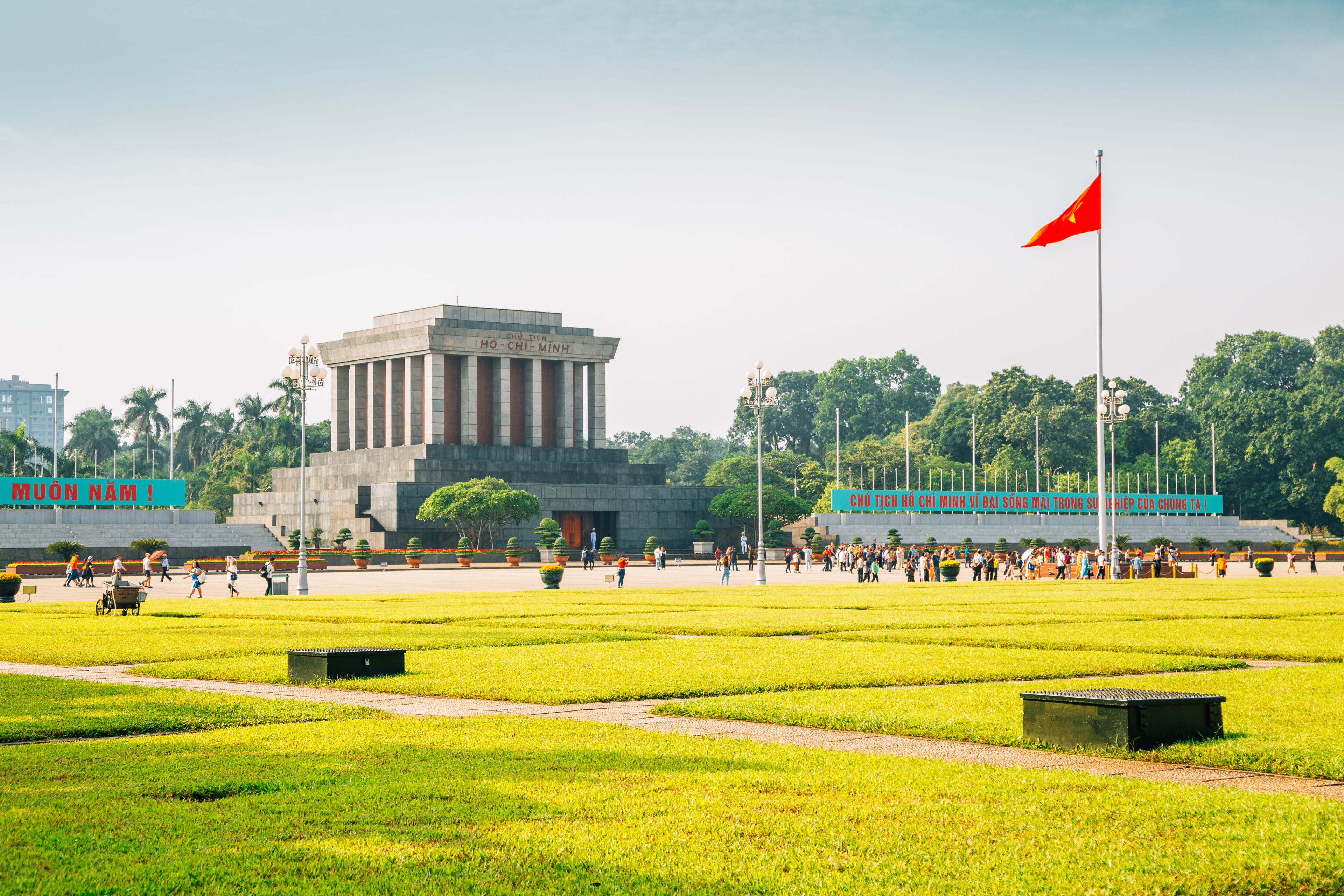 Um edifício grande e histórico com colunas, uma bandeira vermelha e um parque bem cuidado com caminhos.