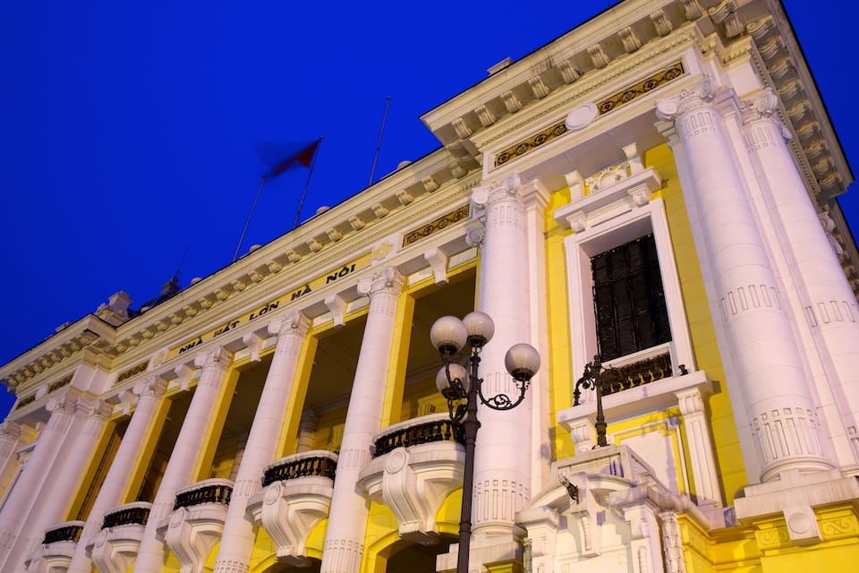Um edifício clássico com colunas brancas e detalhes em amarelo, apresentando decorações elaboradas e uma varanda com balaustrada.