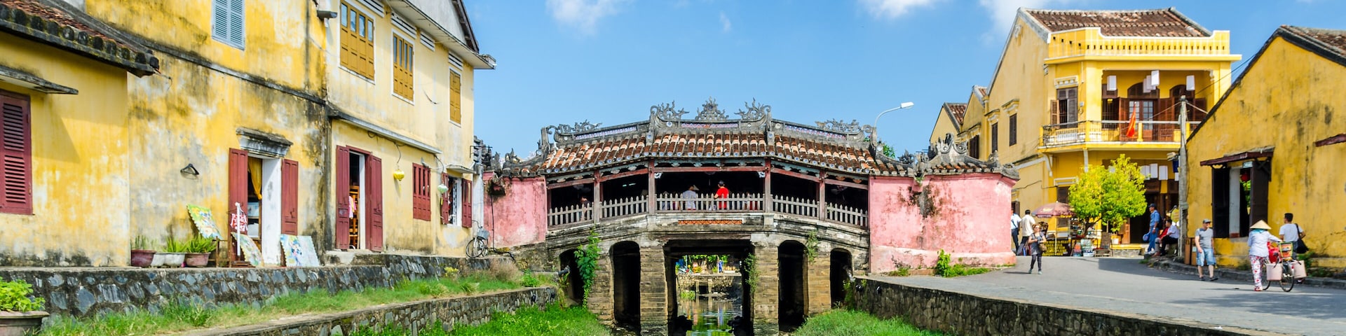 Uma ponte tradicional com grades ornamentadas, um canal refletindo os edifícios e um céu azul límpido.