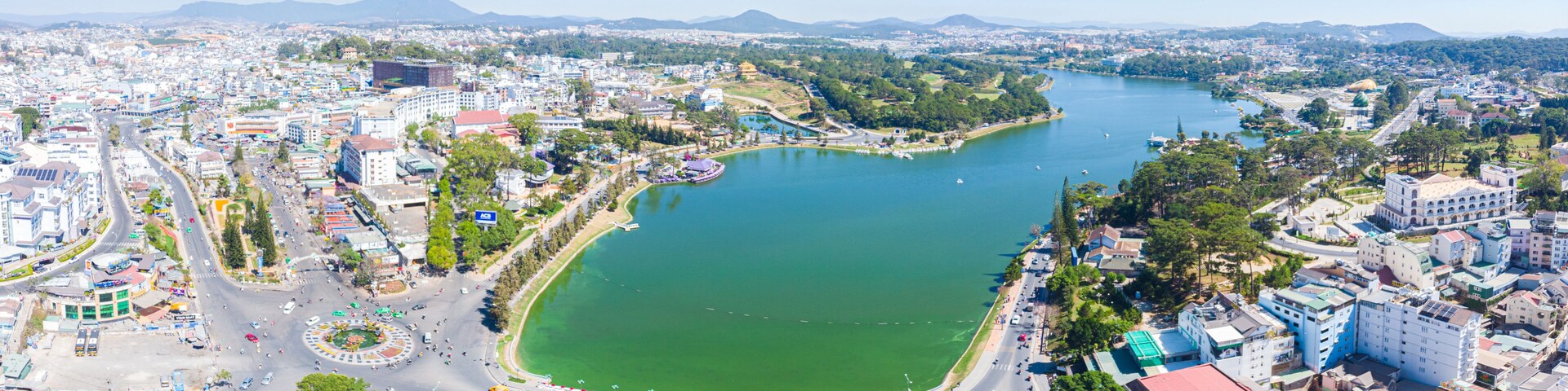 Paisagem urbana com um lago verde grande, edifícios ao redor e montanhas ao fundo.