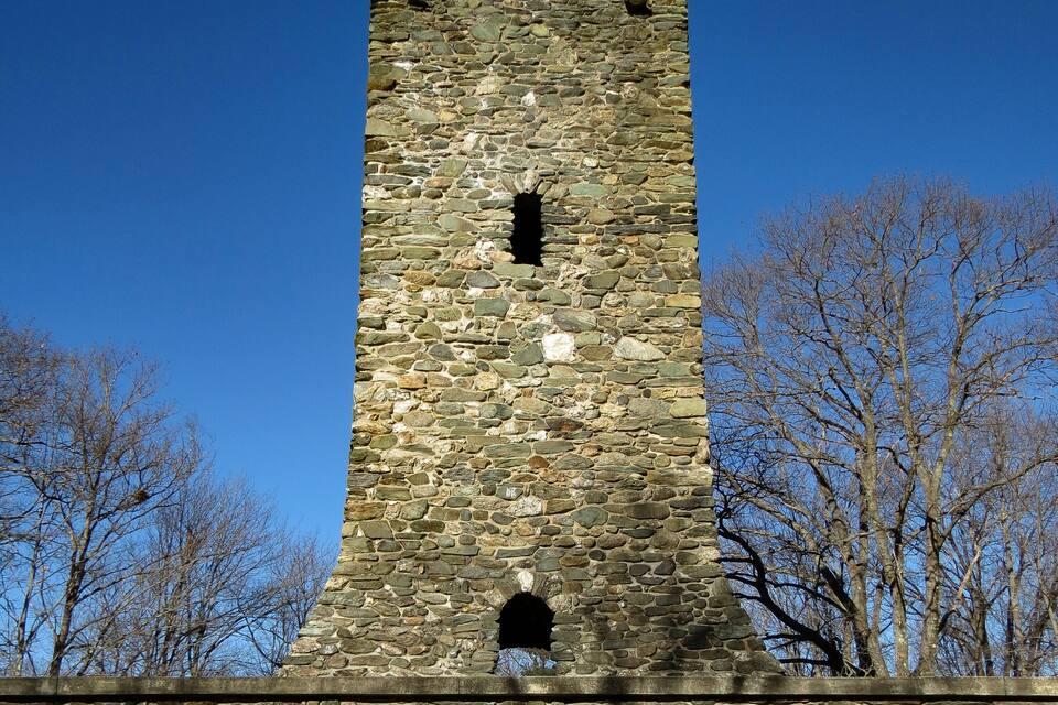 Uma torre de pedra alta com duas aberturas em arco, cercada por grama e árvores.