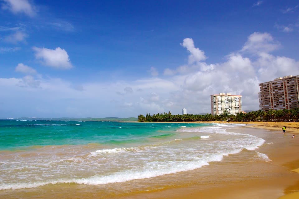 Uma praia com águas azul-turquesa, areal arenoso e edifícios altos ao fundo.