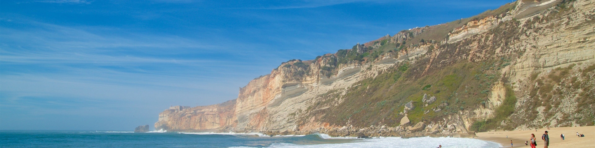 Uma praia arenosa com ondas quebrando na costa, um penhasco rochoso ao fundo e um céu azul límpido.