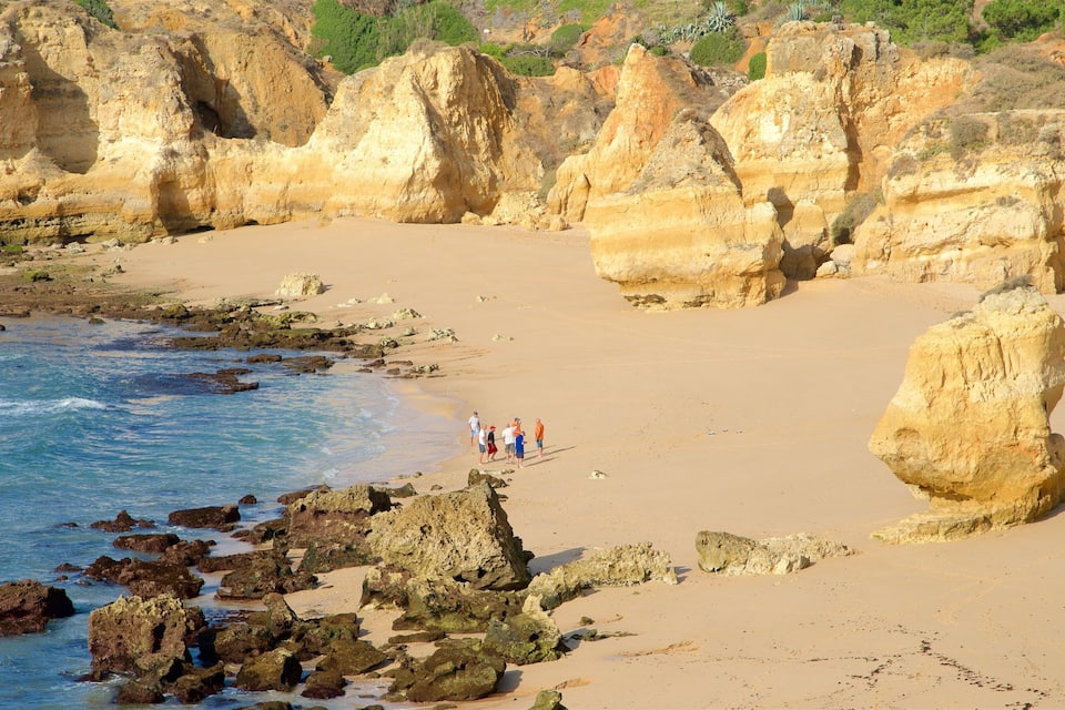 Paisagem costeira com uma praia de areia, falésias rochosas e um grupo de pessoas perto da água.