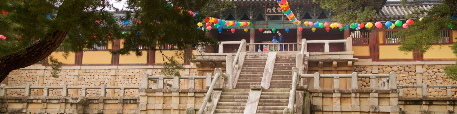 Um edifício tradicional coreano com uma escadaria de pedra que leva até ele, rodeado por árvores e lanternas coloridas.