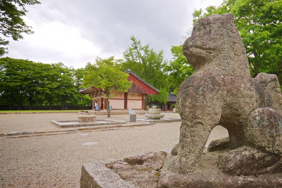 Uma estátua de leão de pedra em um parque, com um edifício tradicional ao fundo.