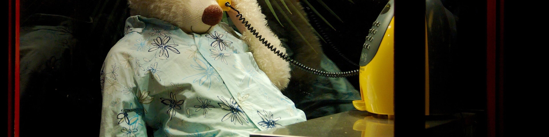 Um ursinho de pelúcia vestindo uma camisa florida sentado à mesa com um telefone amarelo.