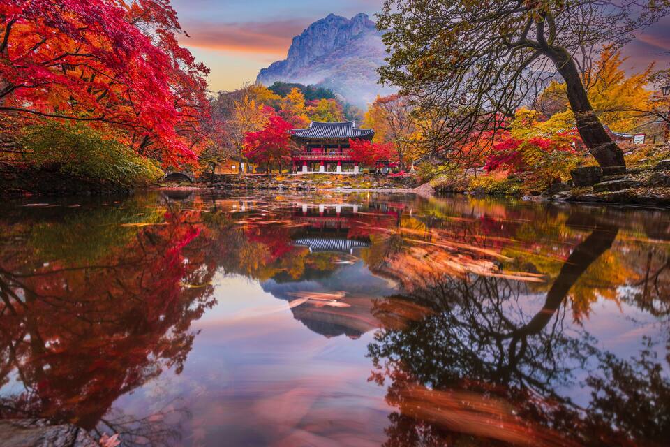 Um pavilhão tradicional junto a um lago espelhado, cercado por folhagem outonal vibrante e com montanhas ao fundo.