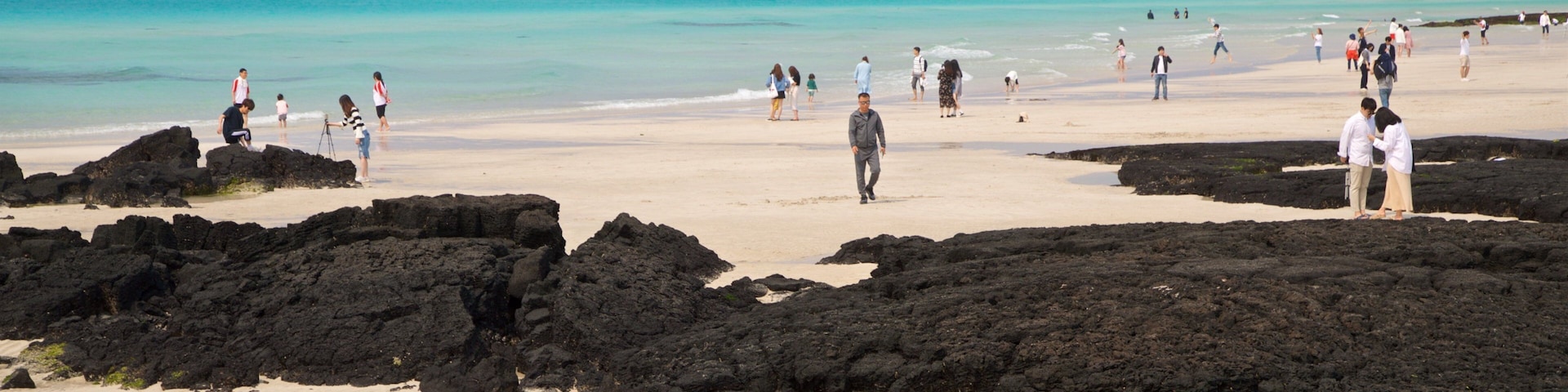 Uma praia com pessoas, um farol e um litoral rochoso.
