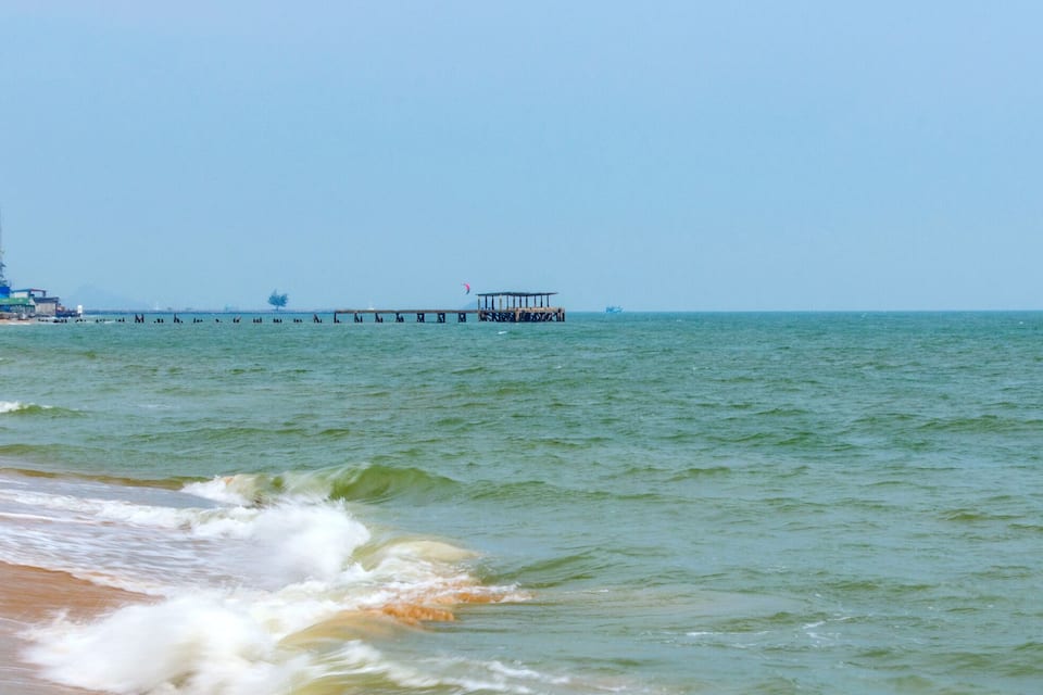 Uma praia com ondas, um cais e edifícios ao fundo.