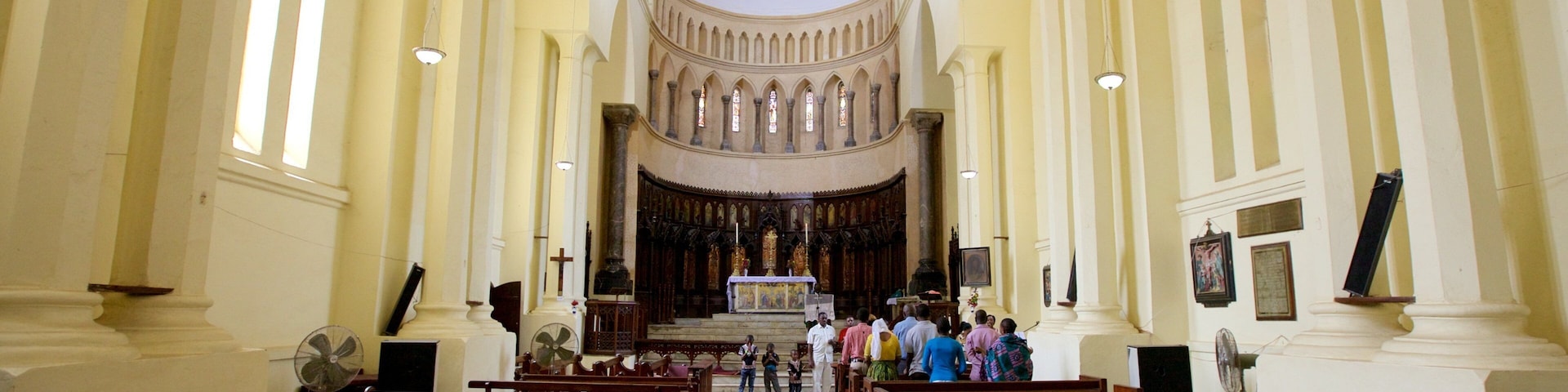 Interior de igreja com fileiras de bancos de madeira, um corredor central que leva a um altar e janelas de vitrais.