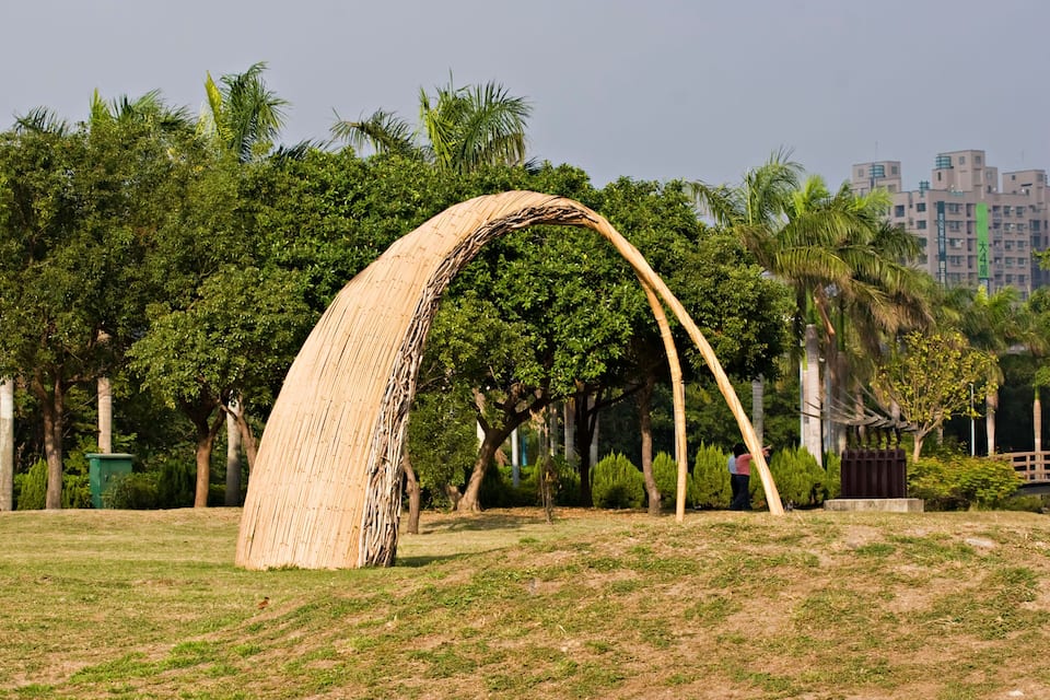 Uma grande estrutura curva de bambu em um parque, com árvores e edifícios ao fundo.
