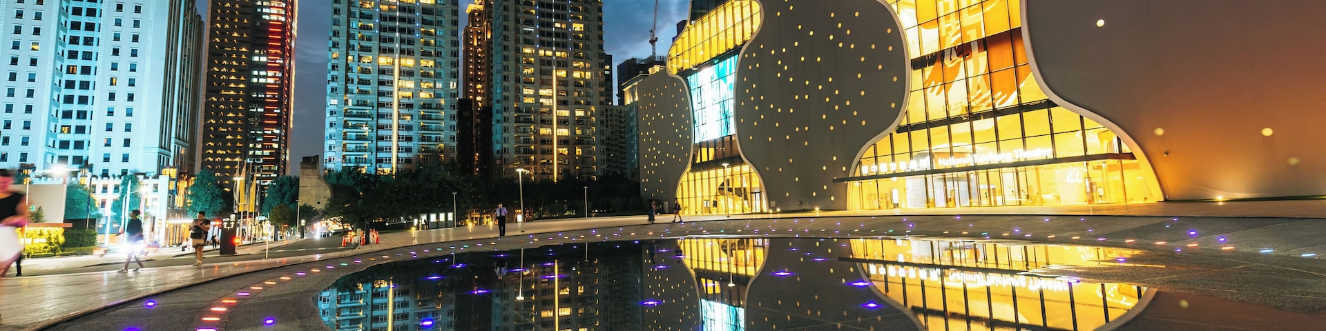 Paisagem urbana moderna com edifícios iluminados e um espelho d'água.