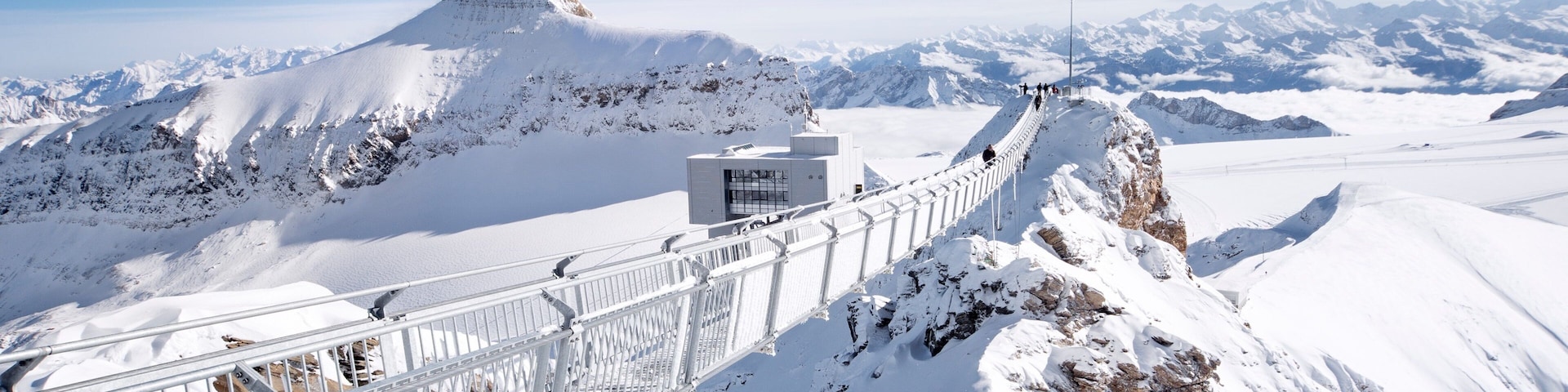 Pico de montanha coberto de neve com plataforma de observação e guarda-corrimão de metal, com vista para uma vasta paisagem nevada.