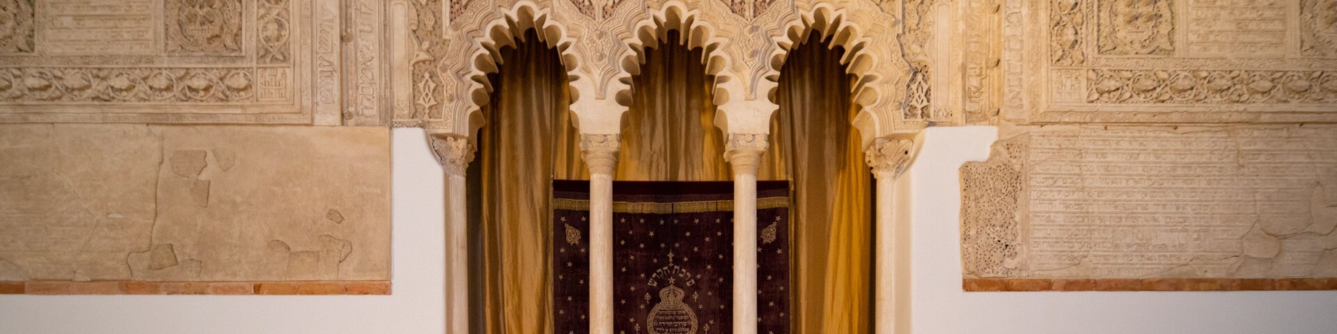 Detalhe arquitetônico ornamentado com esculturas elaboradas e uma janela com três colunas e cortinas drapeadas.