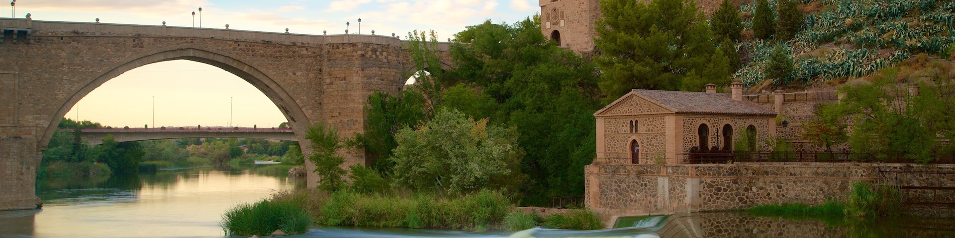 Uma ponte de pedra sobre um rio, um castelo e vegetação exuberante.