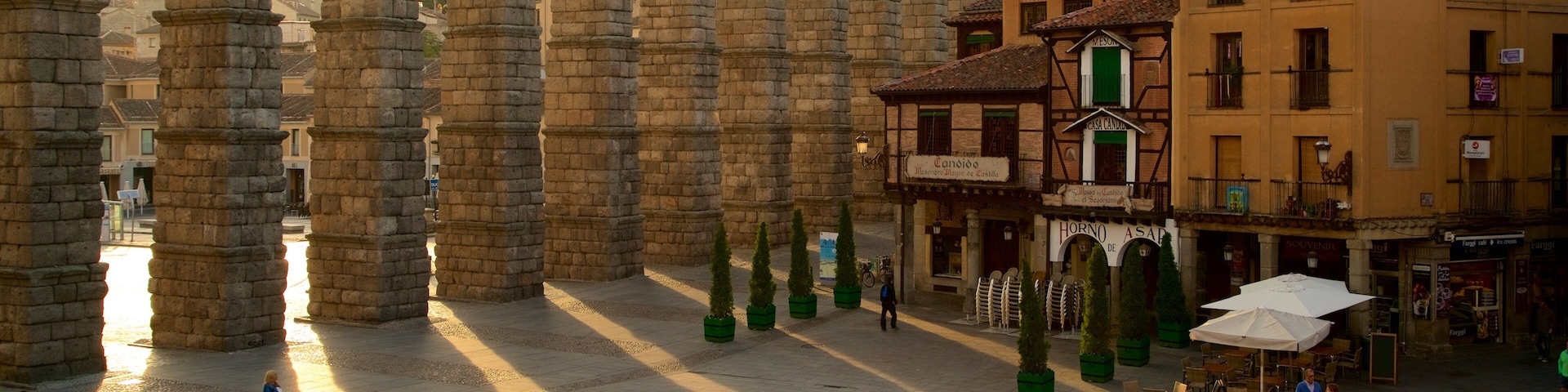 Um antigo aqueduto com arcos, uma praça com pessoas a caminhar e edifícios tradicionais com placas.