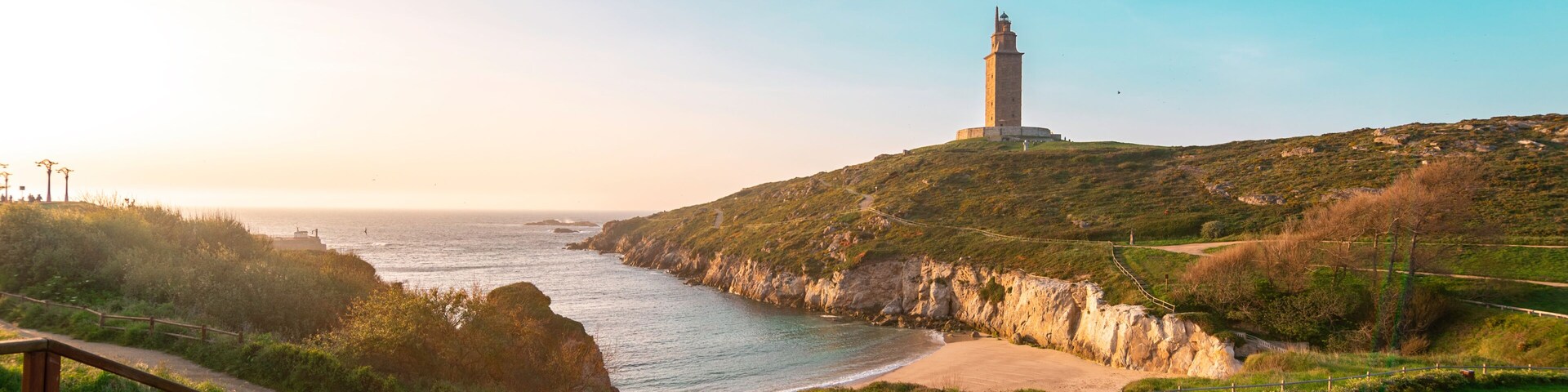 Paisagem costeira com um farol, colinas gramadas, uma praia e uma escadaria de madeira que leva ao farol.