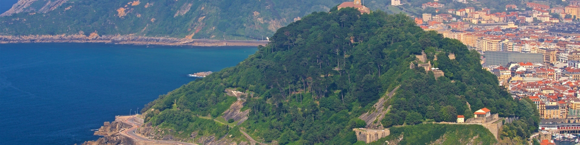 Uma cidade costeira com uma fortaleza imponente no topo de uma colina e uma estrada sinuosa ao longo da costa.
