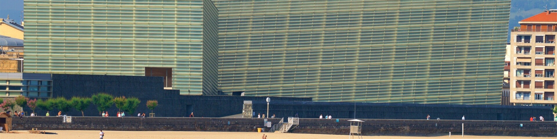 Uma praia com pessoas tomando sol e nadando, e um edifício moderno grande com fachada em grade.