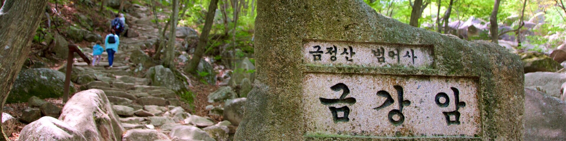 Placa de pedra com texto coreano numa área arborizada com trilho pedregoso.