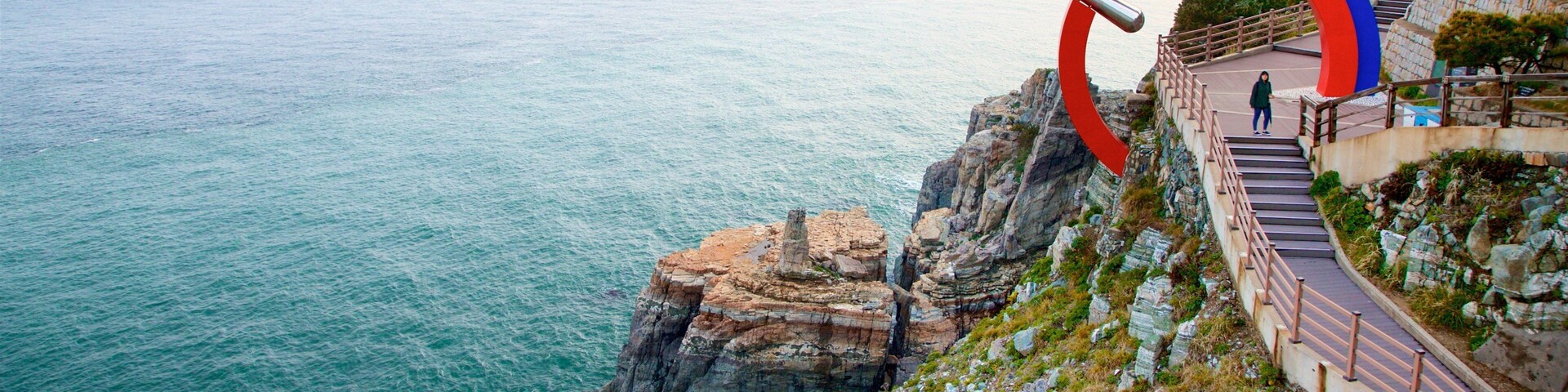 Um penhasco costeiro com uma escultura de metal, uma escadaria e vista para o mar.